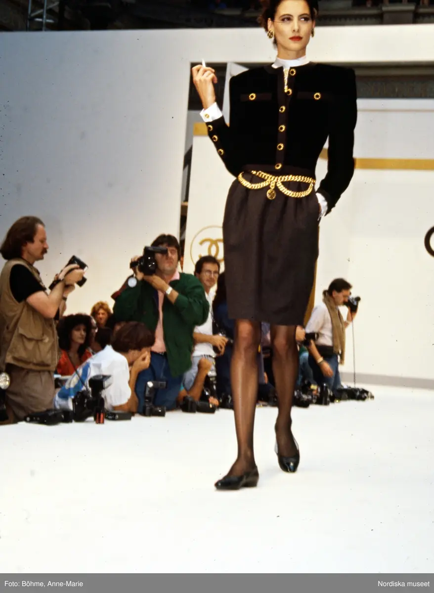 Modevisning. Modell i svart jacka med guldknappar, kjol och guldkedja ...