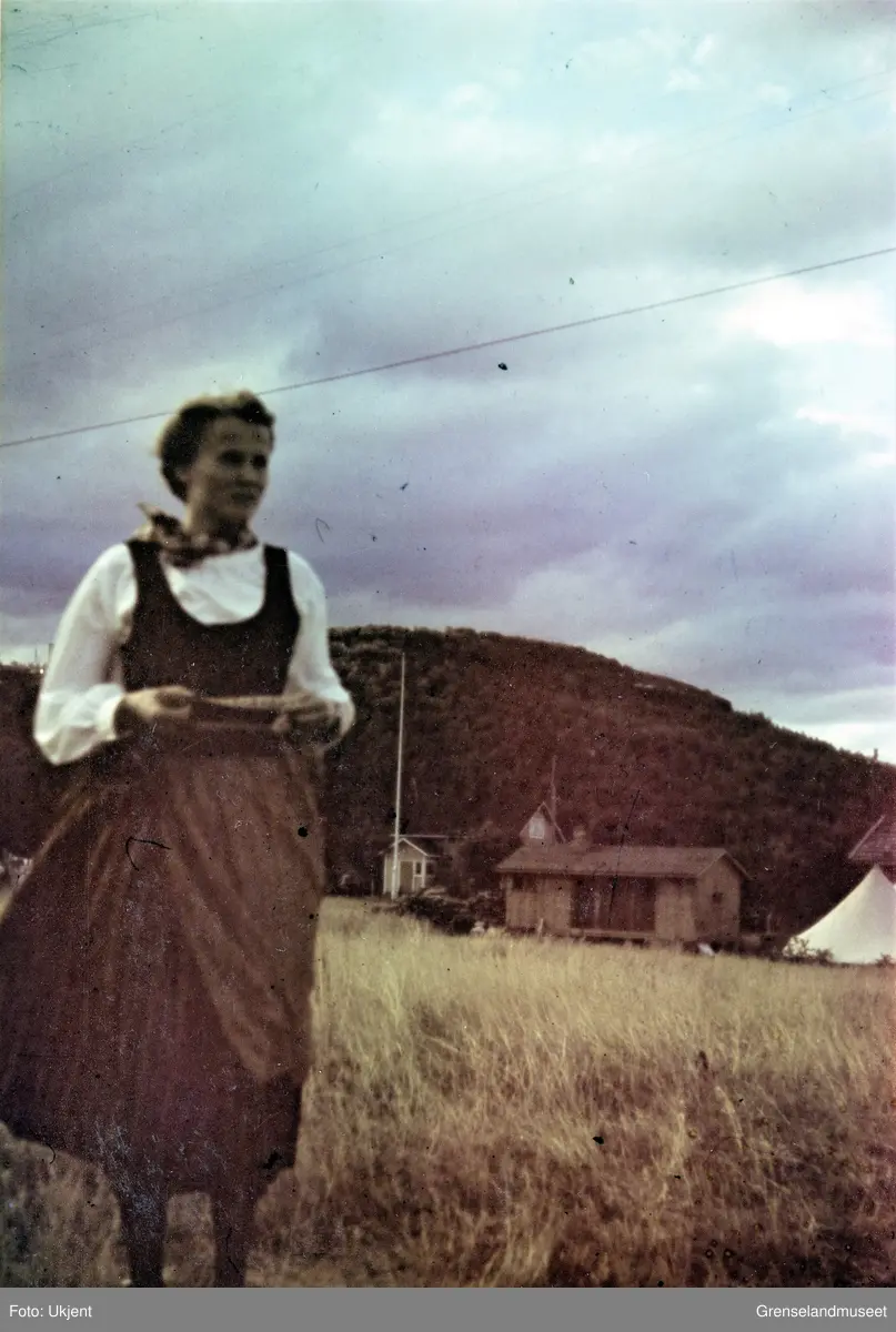 En kvinne står ute på et jorde eller tun. I bakgrunnen står noen ...