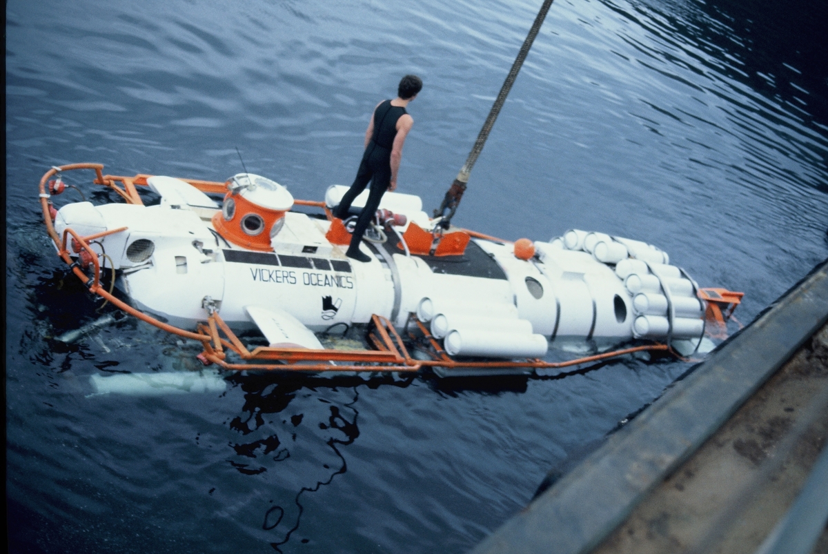 En ubåt løftes fra en lektert og ut i sjøen. En mann i våtdrakt står ...