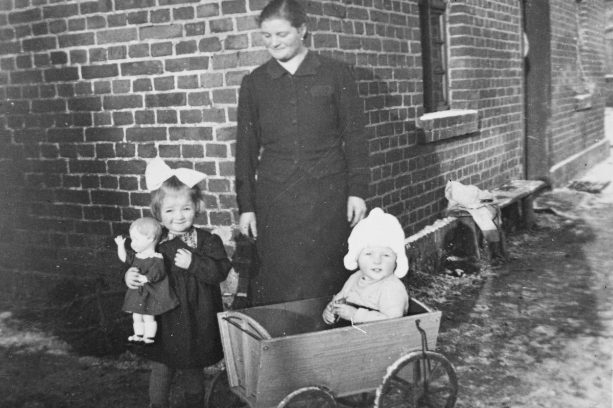 To små barn og en voksen dame ved en murbygning. Det ene barnet har ...